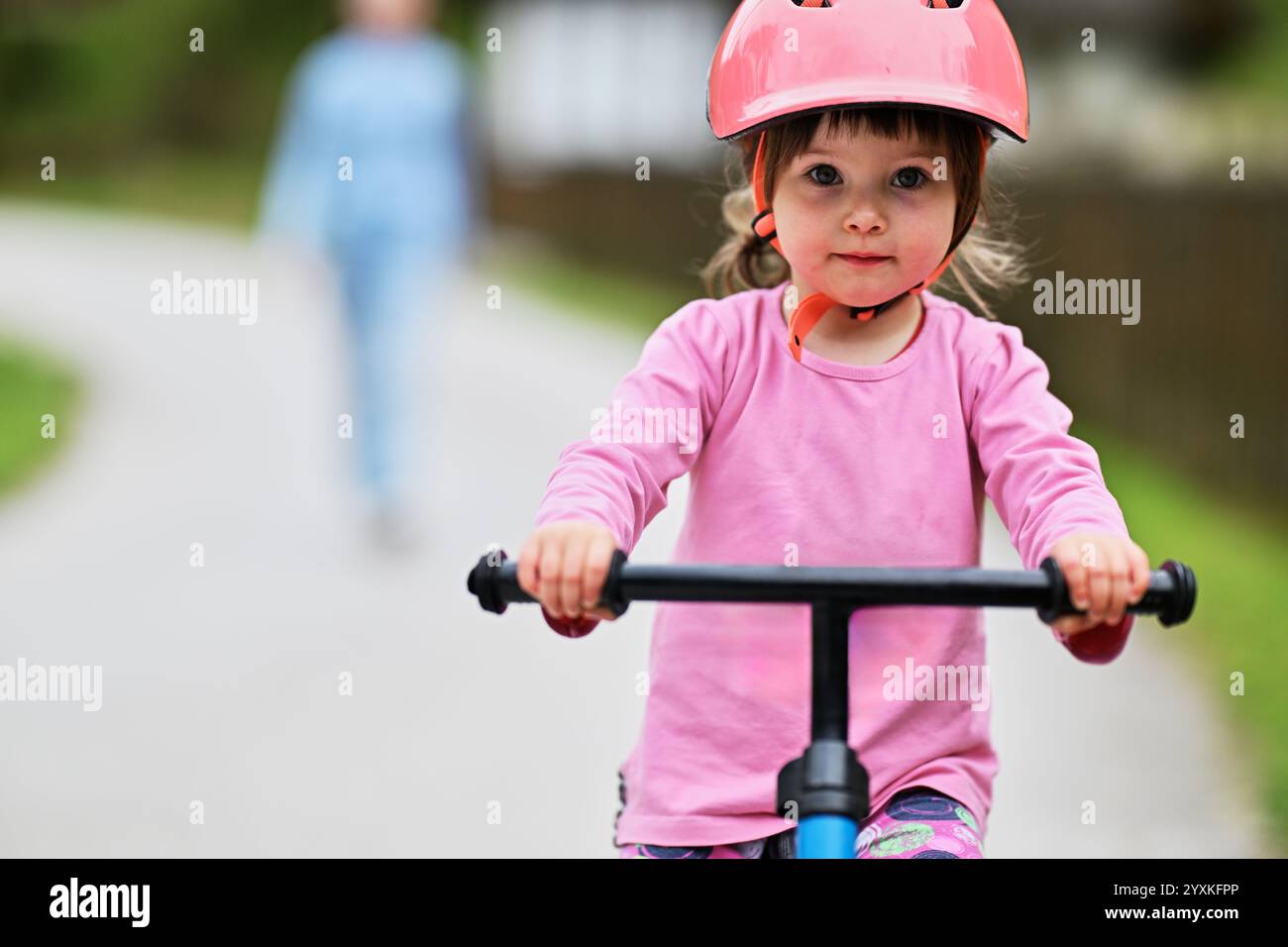 A little girl, dressed in a cute outfit and wearing a helmet, excitedly learning to ride her ...