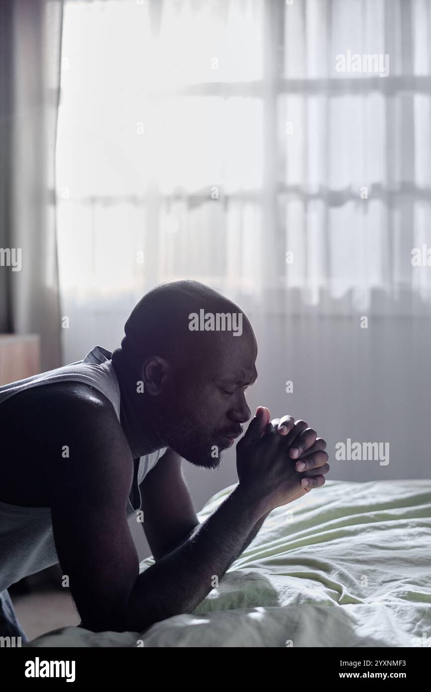 Vertical shot of African American man kneeling while praying on bed ...