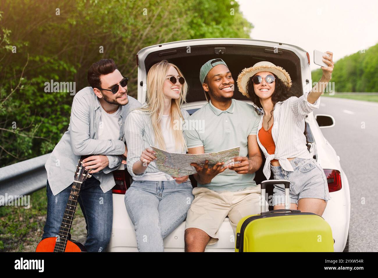 Group of best friends checking map during road trip, sitting in car trunk, taking selfie on ...