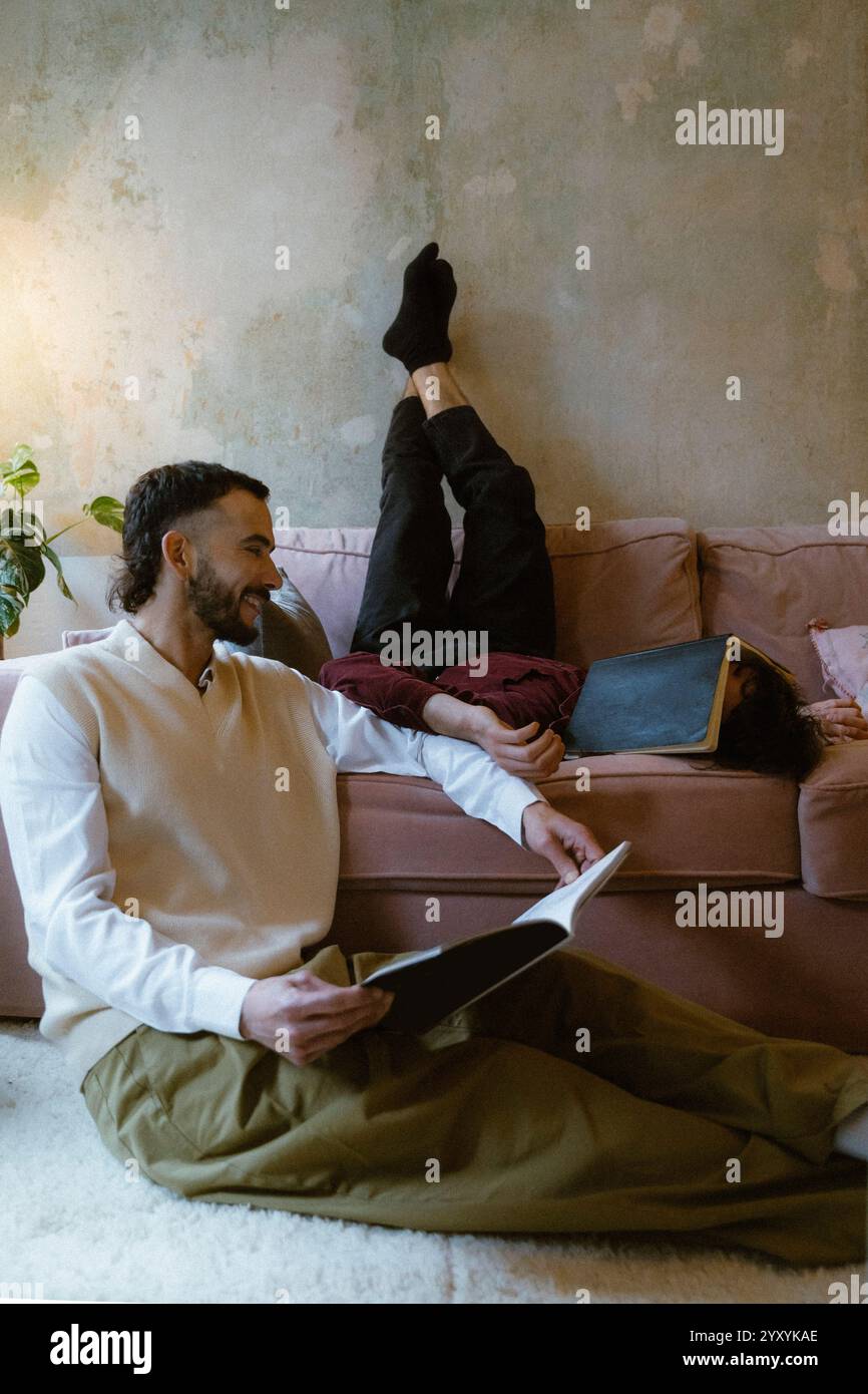 Happy gay man sitting and holding magazine while boyfriend lying down ...