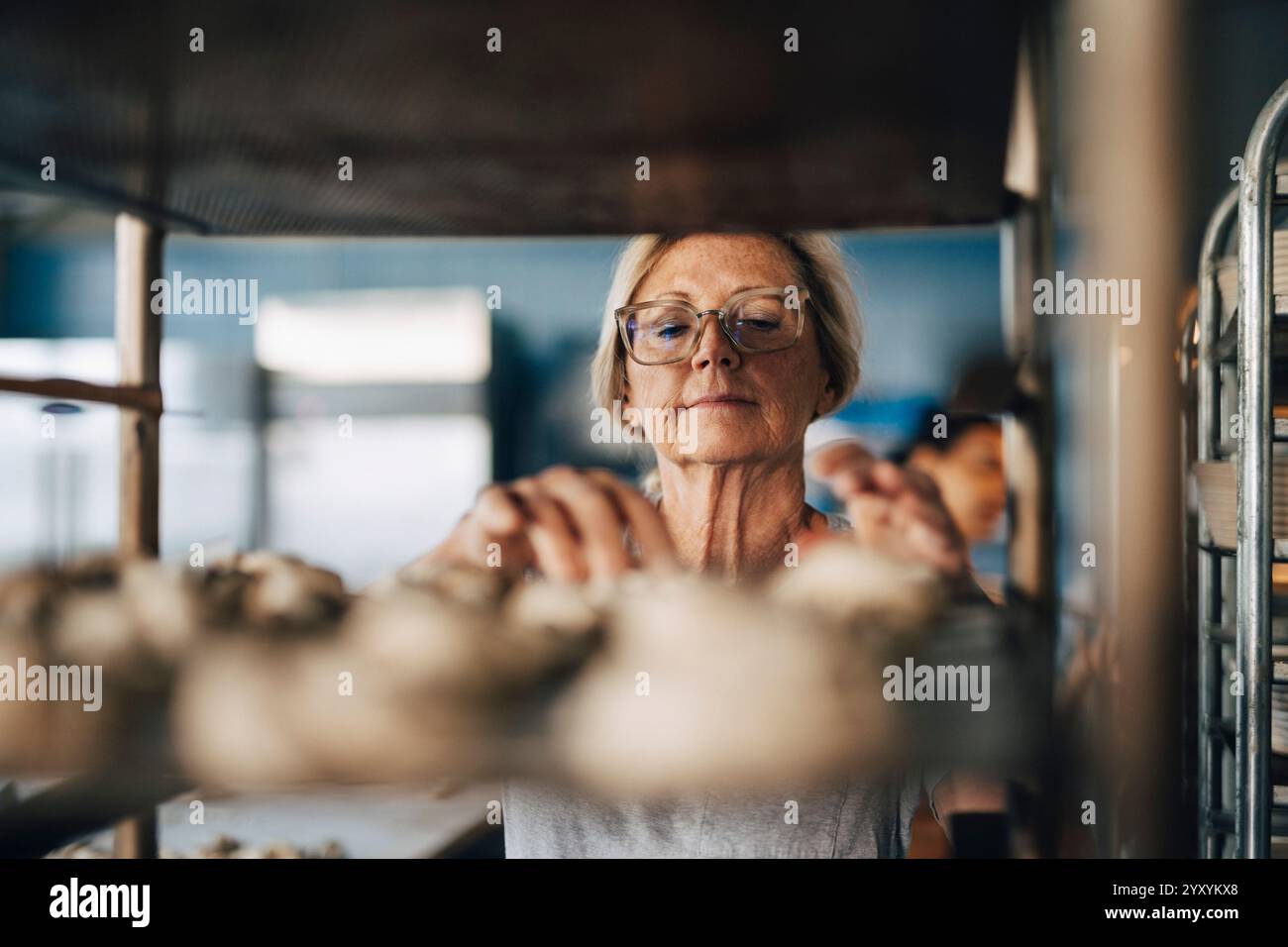 Female senior baker arranging buns on cooling rack while working at ...