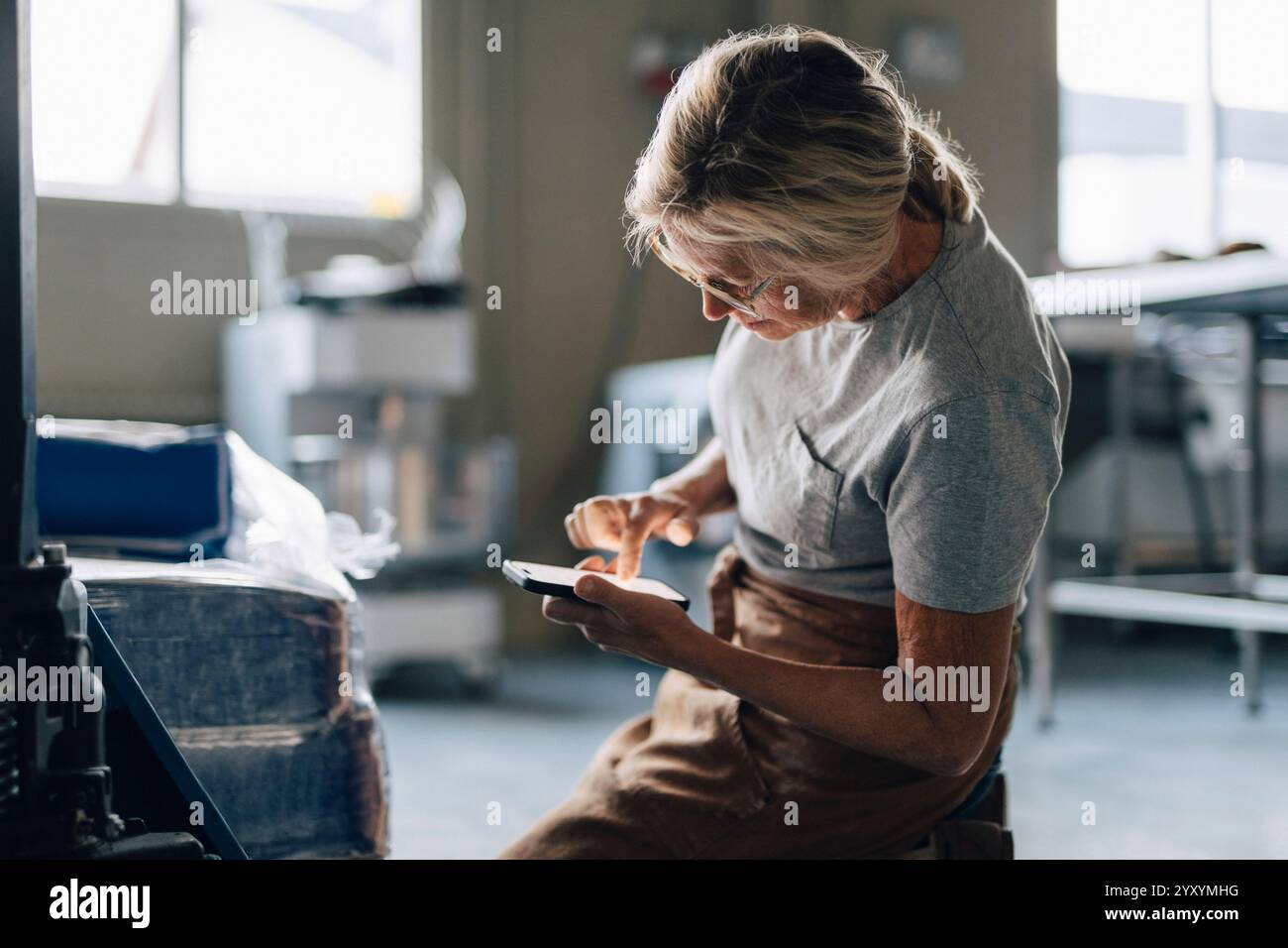 Senior female baker taking inventory of flour sacks through smart phone while working at bakery ...