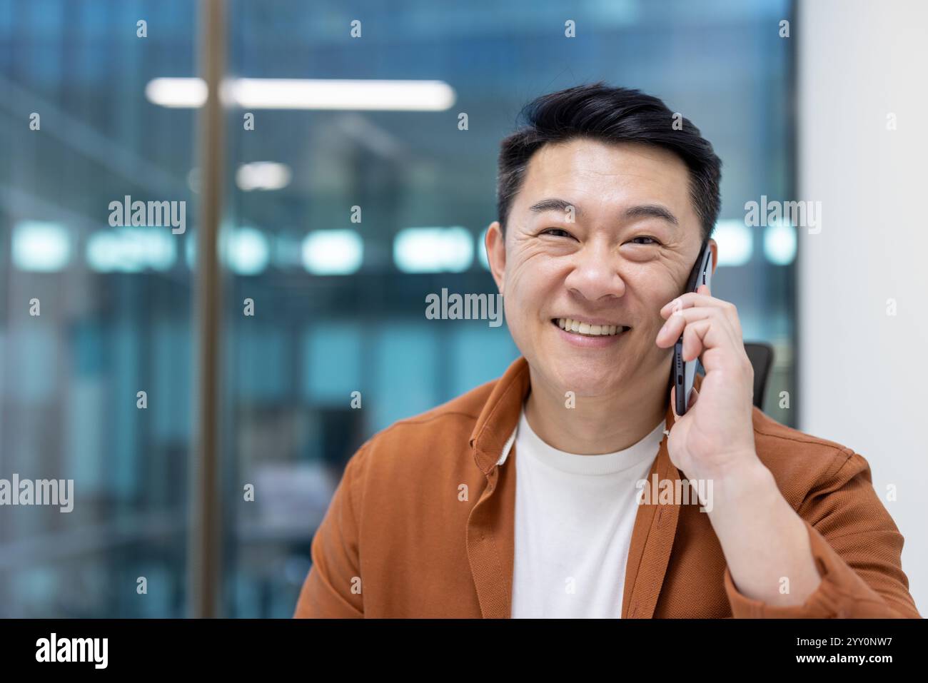 Asian man talking on the phone with a smile, sitting in a bright office ...
