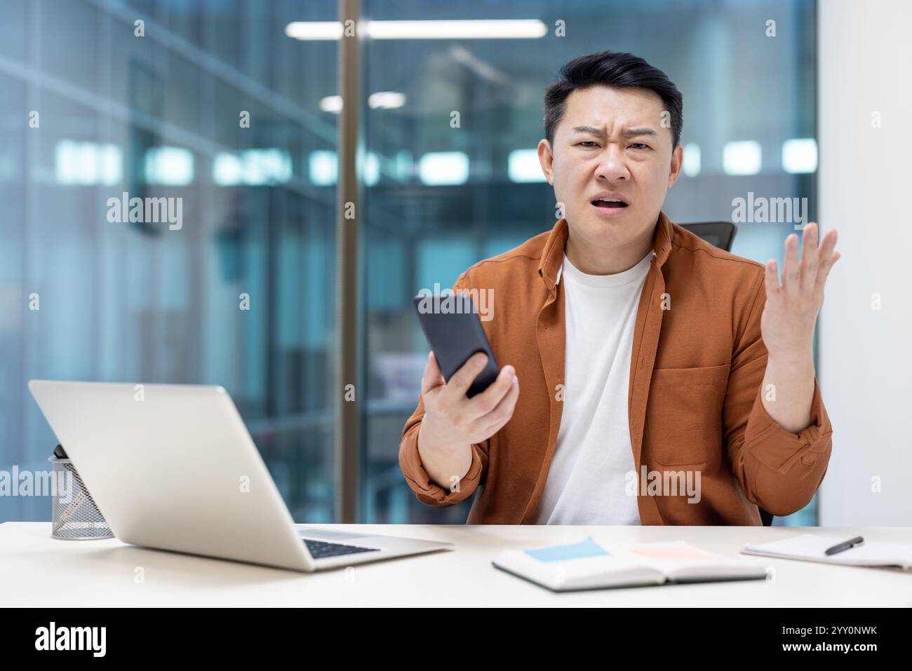 Asian man displaying frustration in a modern office, seated at a desk ...