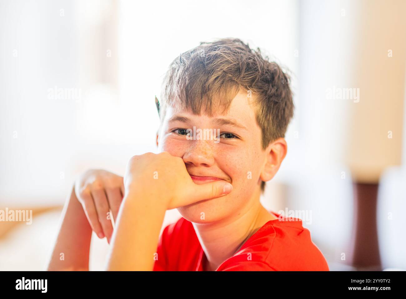 A young boy with short hair and a bright red shirt smiles while resting ...