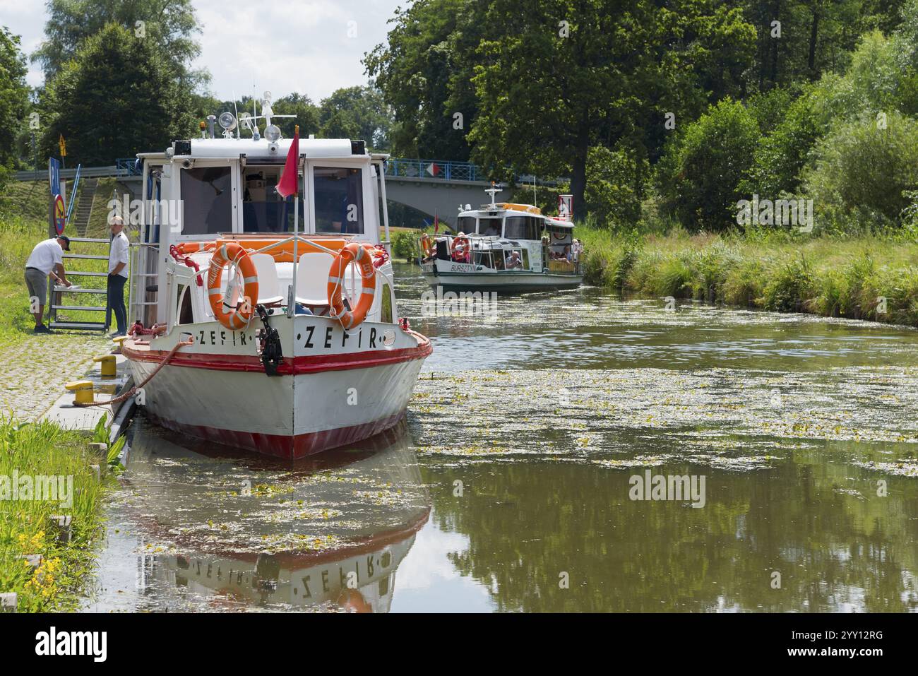 Boats on the bank of an overgrown canal in front of a bridge, people on ...