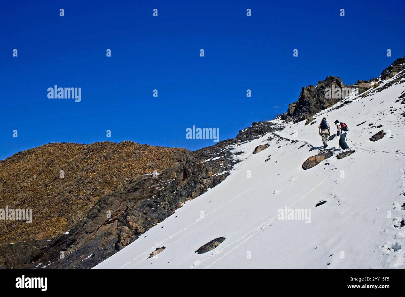 Two hikers ascend a snow-covered slope in Bolivia, surrounded by rugged ...