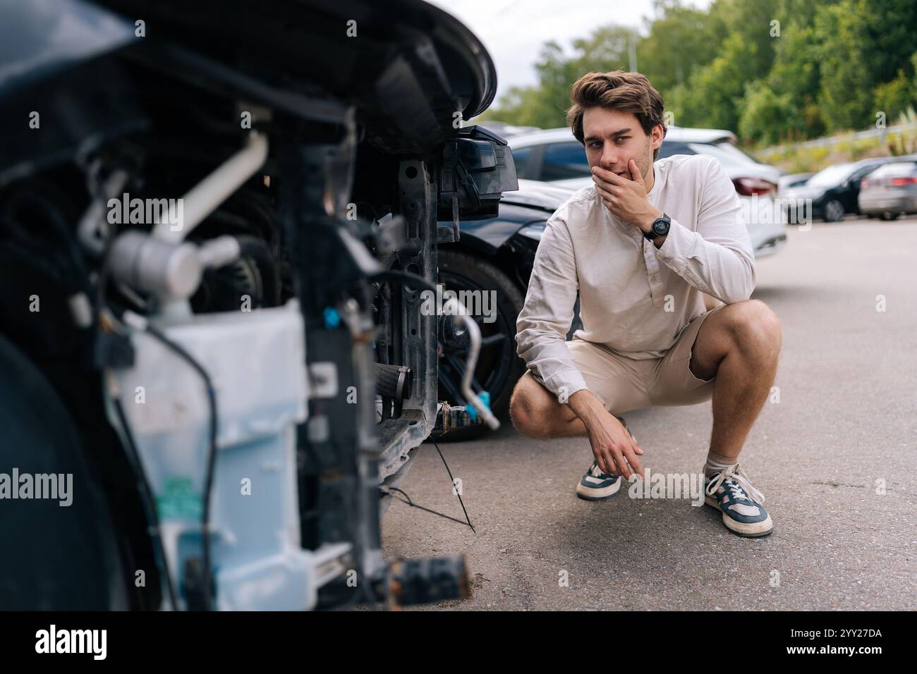 Pensive young driver crouching next to damaged car in parking lot ...