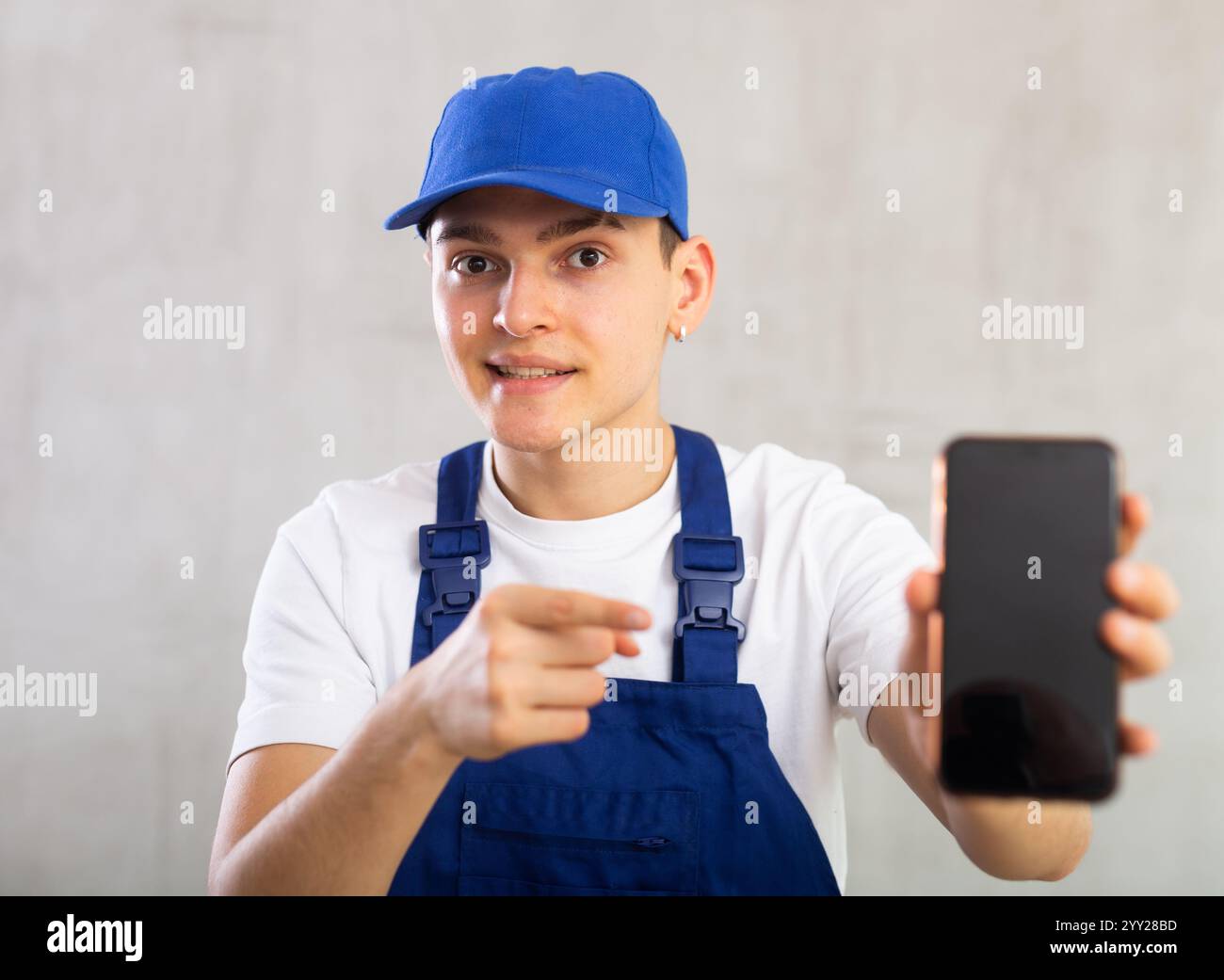 Man in jumpsuit and cap shows dark empty mobile phone screen and points ...