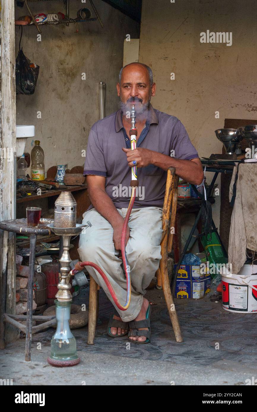 Middle-aged man enjoying a traditional hookah in a rustic Alexandria workshop, surrounded by ...