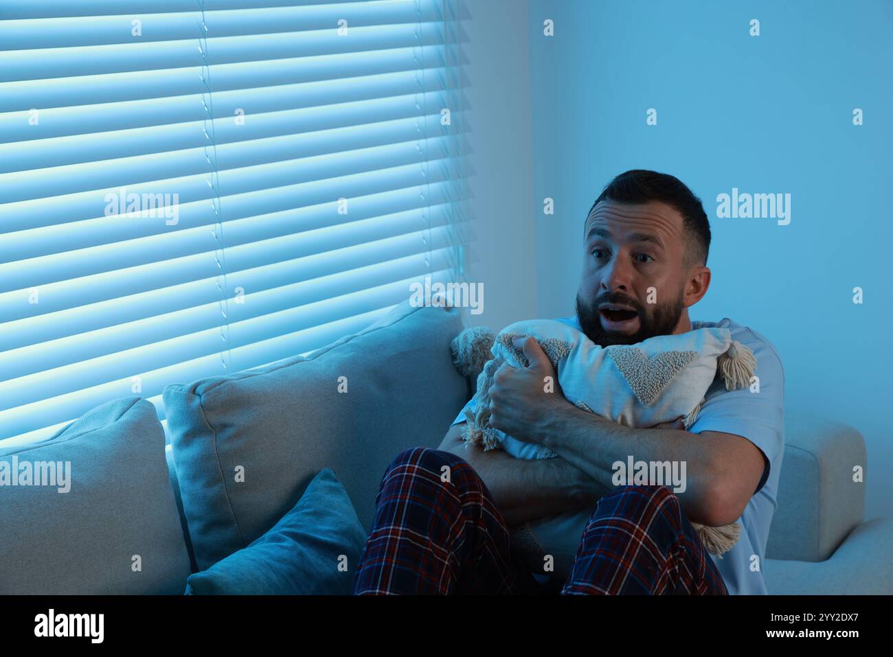 Fear of darkness. Scared man hugging pillow on couch in dark room ...