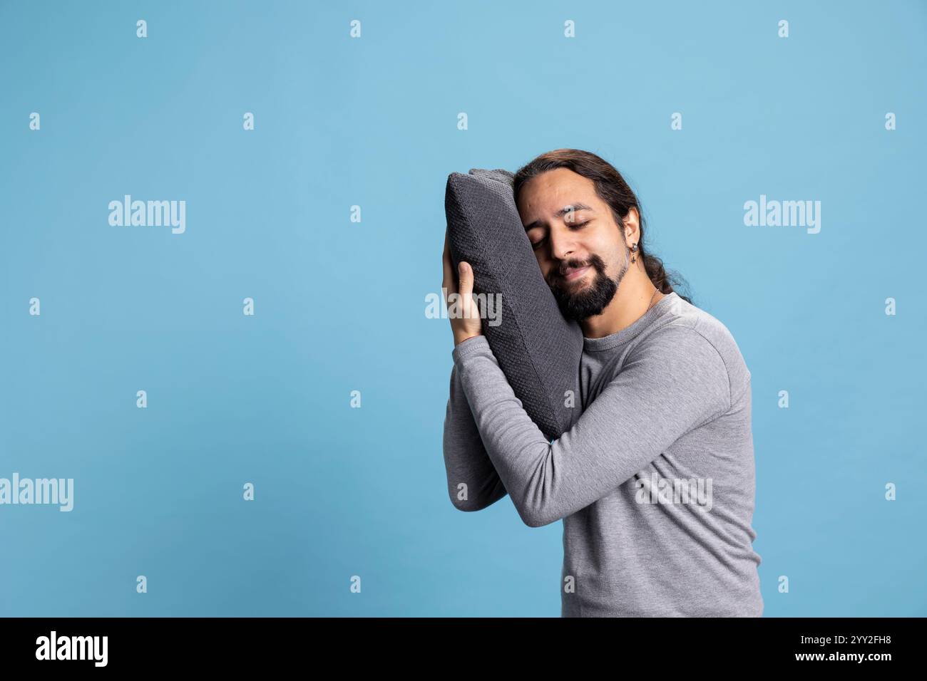 Sleepy young man taking a nap with a pillow standing against blue ...