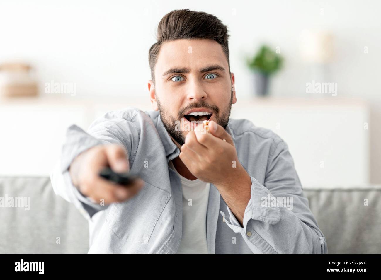 Emotional man eating popcorn and holding remote controller, watching TV ...