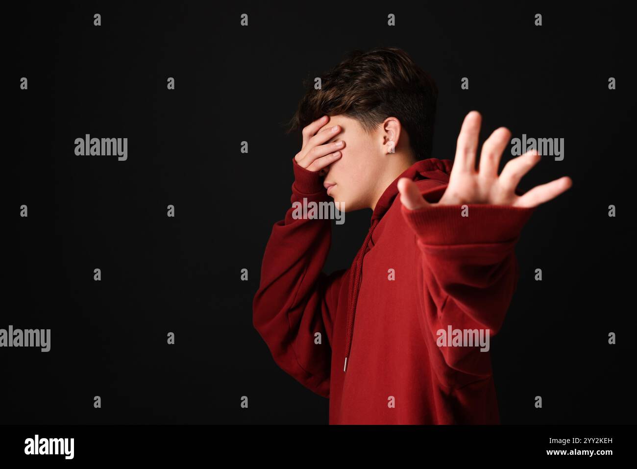 Scared teenage boy covering face with hands on black background. Space ...
