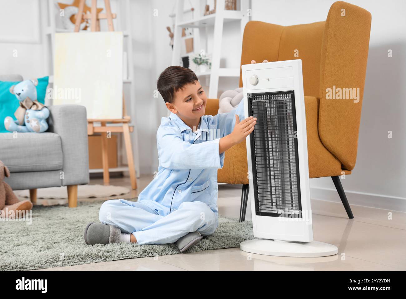 Cute boy in pajamas with electric heater sitting on carpet at home ...