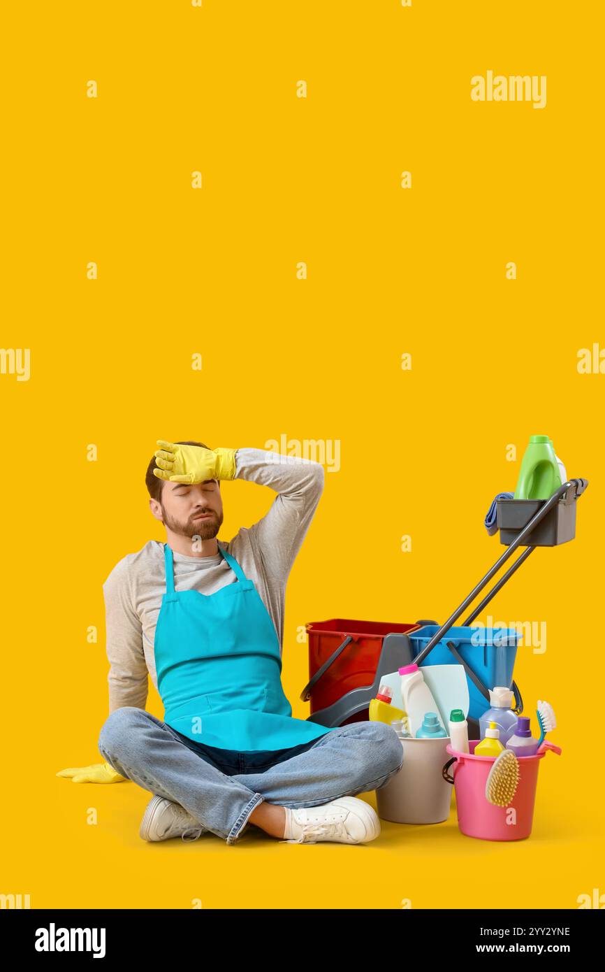 Tired male janitor with cleaning supplies sitting on yellow background ...