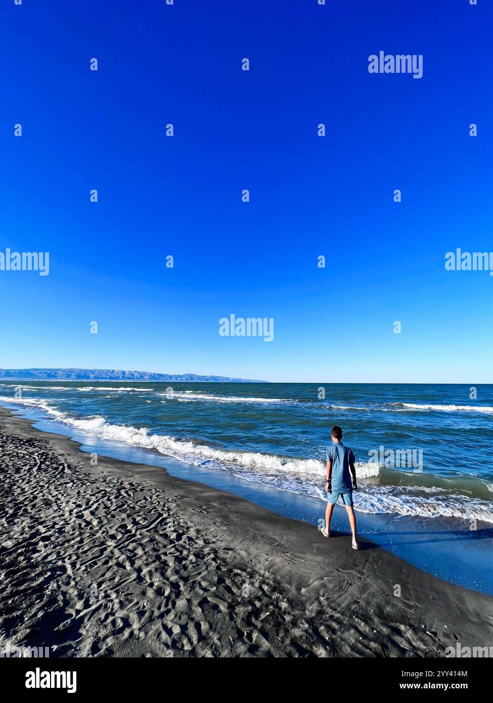 A boy strolls along a sandy beach, enjoying the serene seaside ...