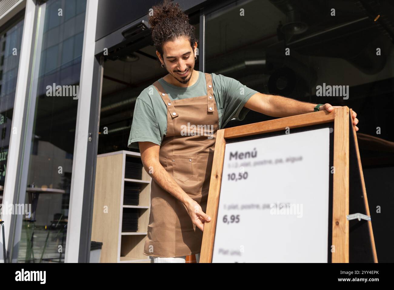 A cheerful cafe worker in a brown apron sets up a wooden menu board ...