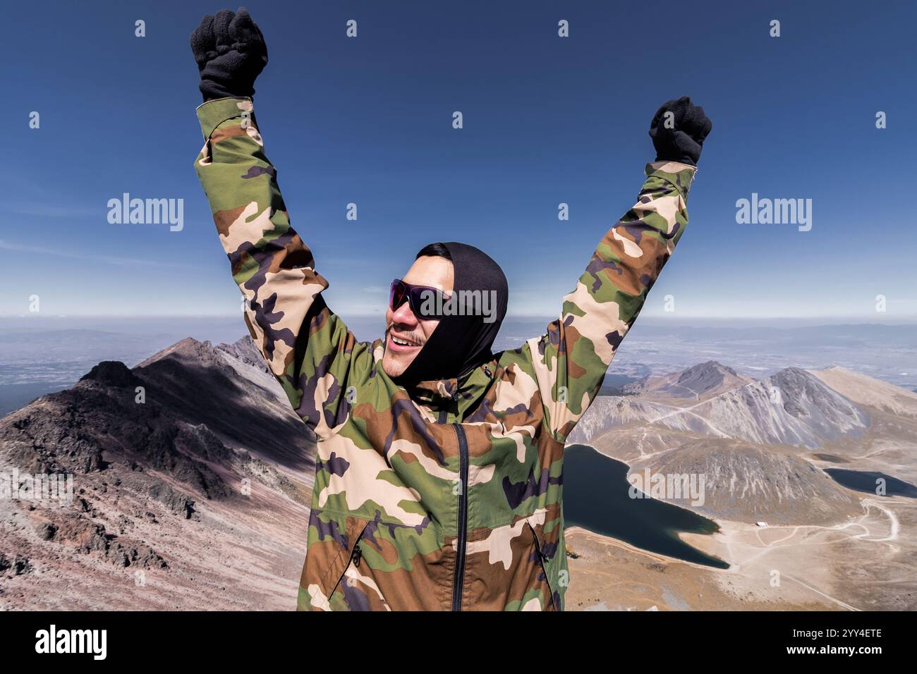 A hiker in a camouflage jacket celebrates triumphantly on a mountain ...