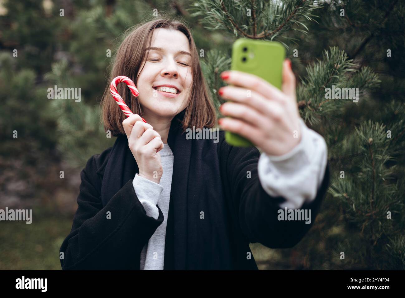 A joyful woman captures a selfie holding a candy cane amidst pine trees ...