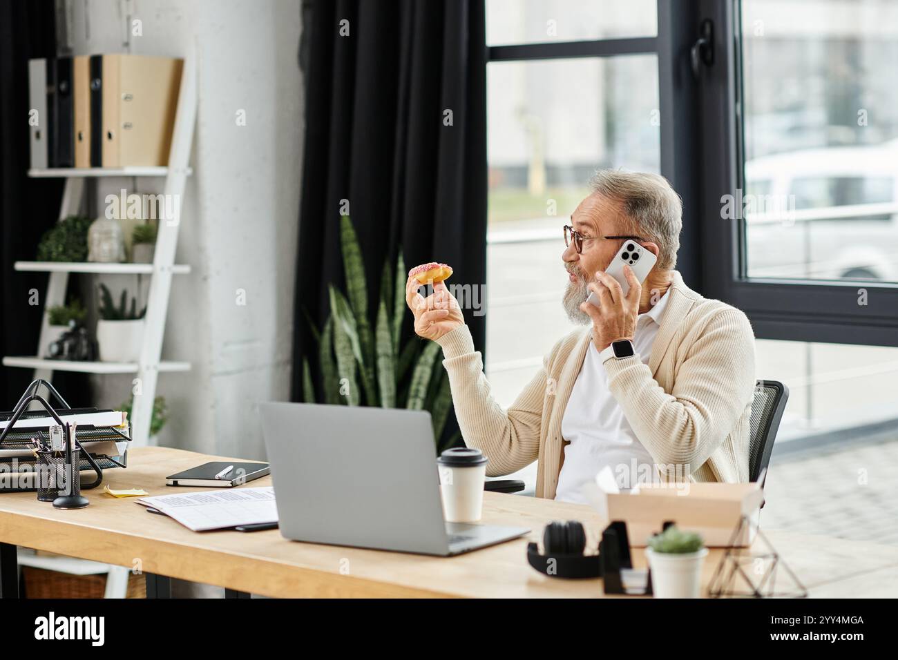Handsome older man multitasks at his desk, enjoying a donut and talking ...