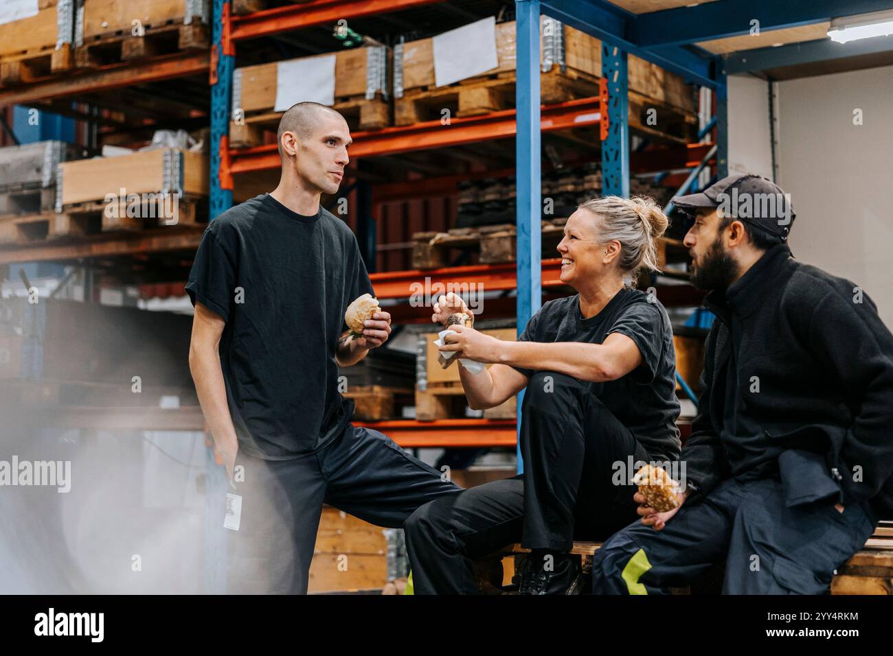 Male and female warehouse workers eating burgers while talking with ...