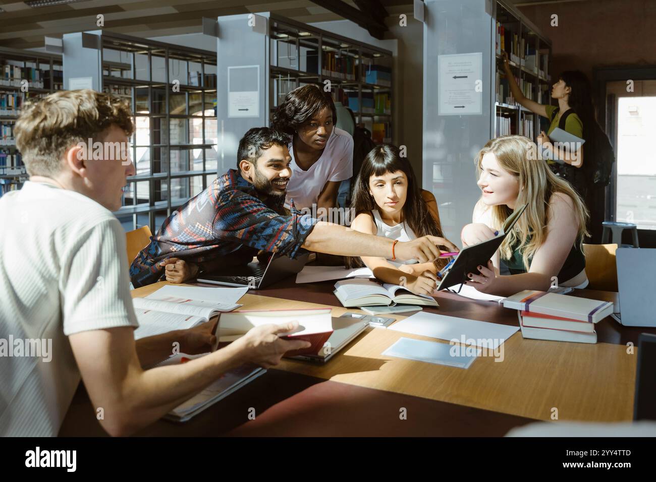 Smiling man pointing at book while doing group study with male and ...