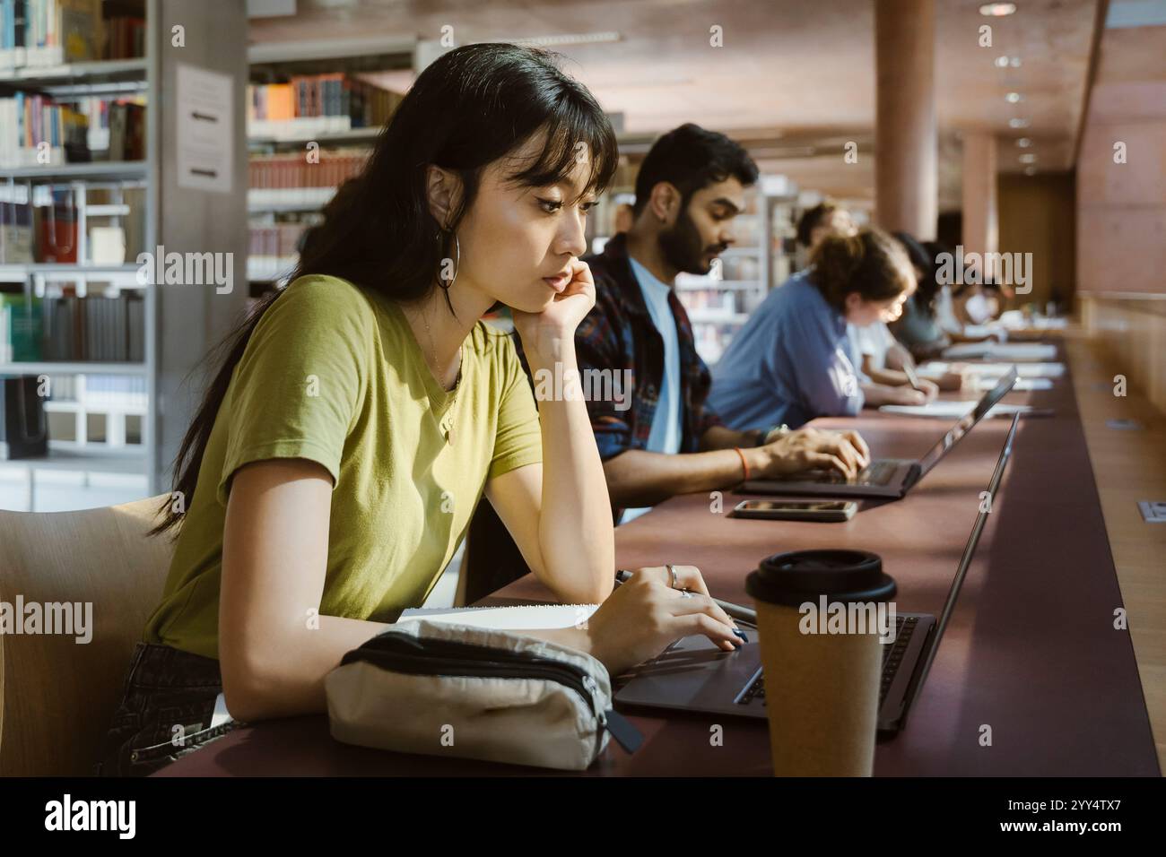 Focused young woman studying on laptop while leaning on elbow at desk in college library Stock ...