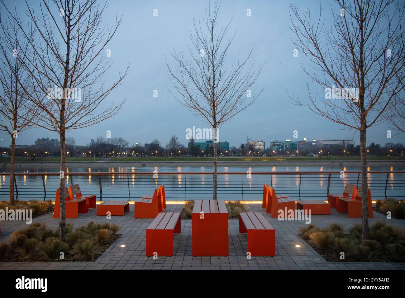 Modern wooden benches and tables on cement floor in city park at evening. River and city lights ...