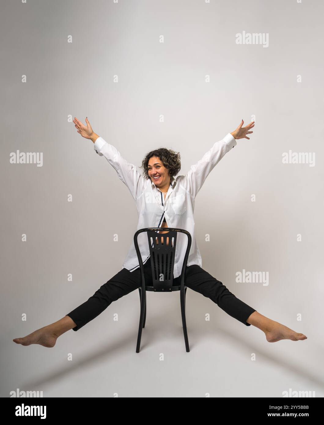 Happy positive woman in white shirt, sitting on chair, arms raised ...