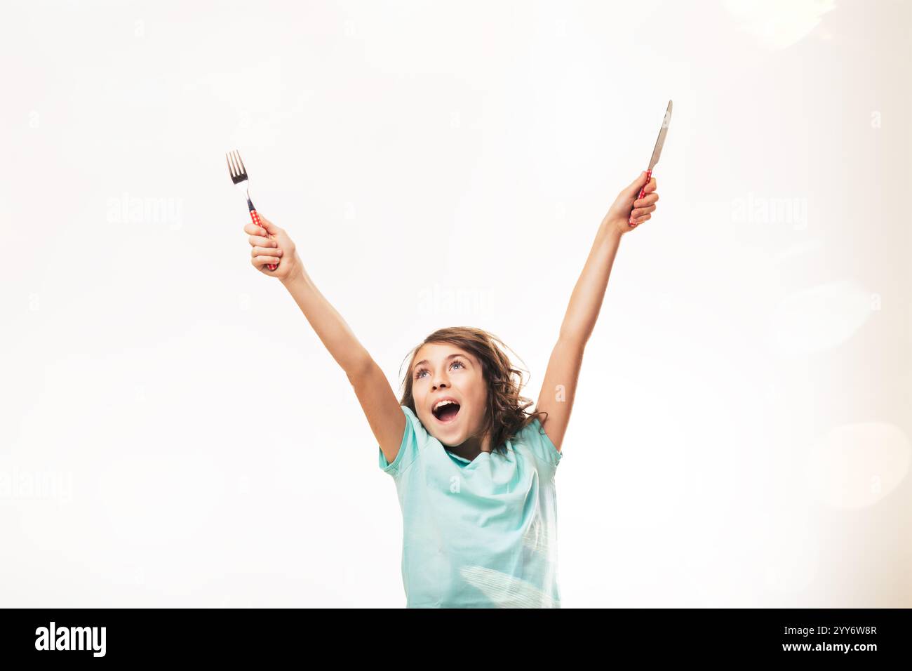 Enthusiastic preteen girl raising her arms, holding a fork and knife ...