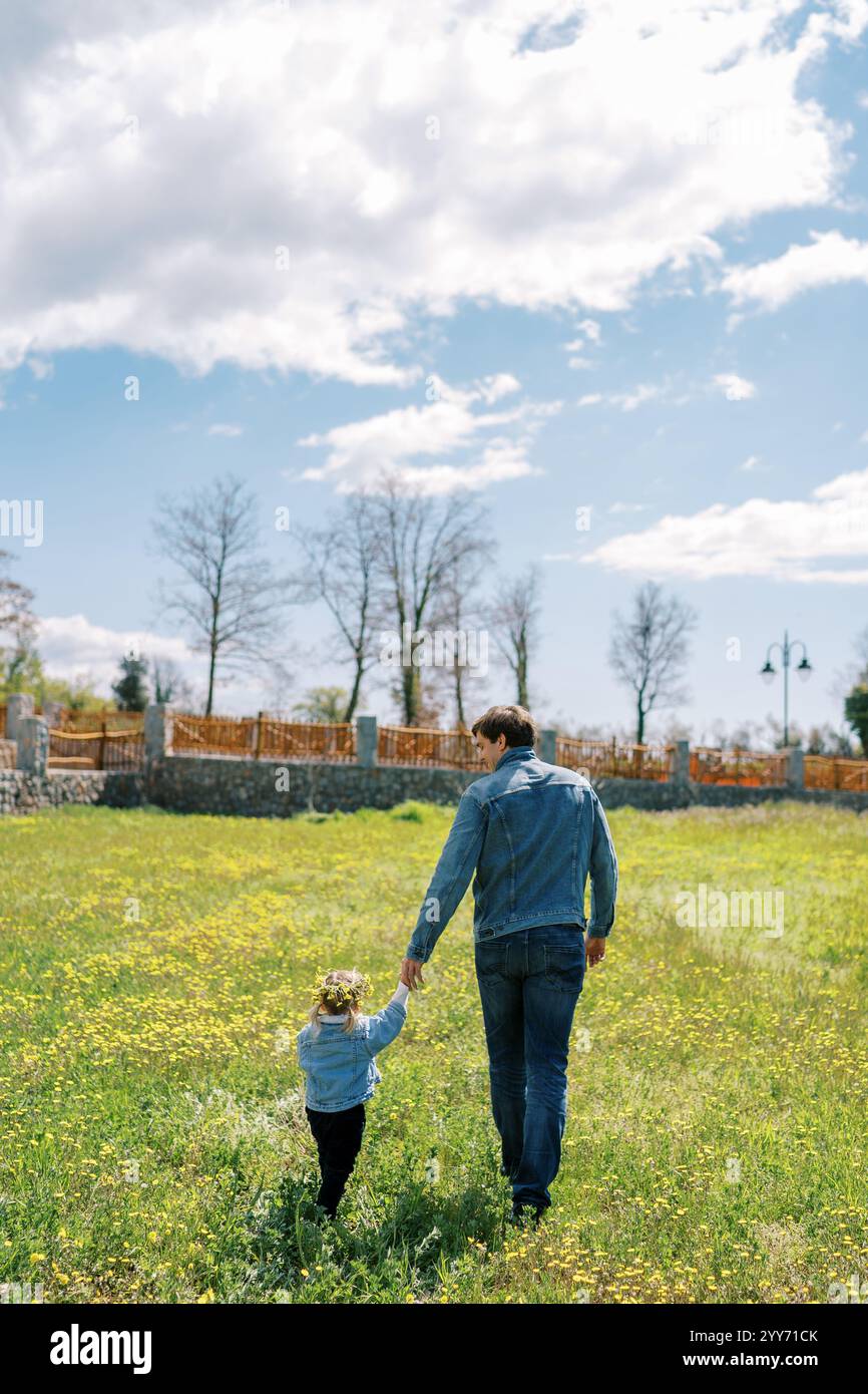 Little girl in a wreath walks through a flowering field with her dad by the hand. Back view ...