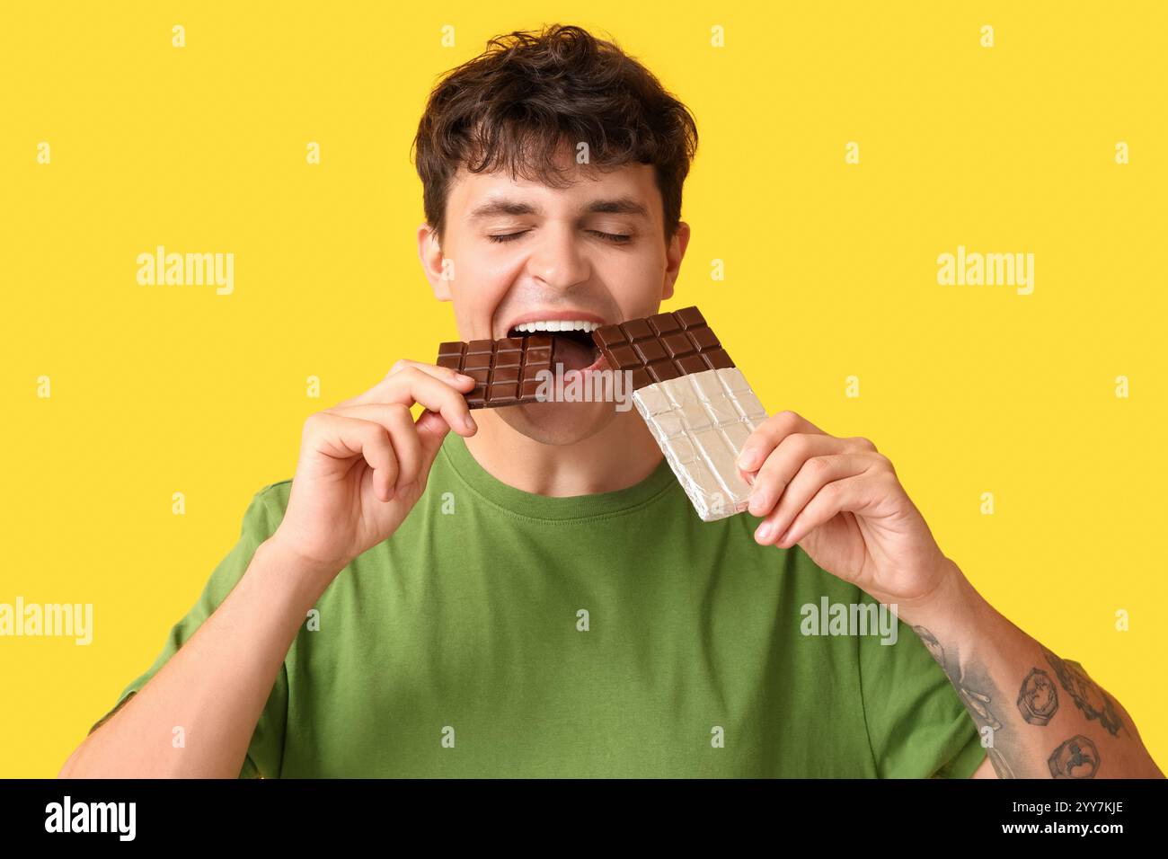 Handsome young man eating sweet chocolate bars on yellow background ...