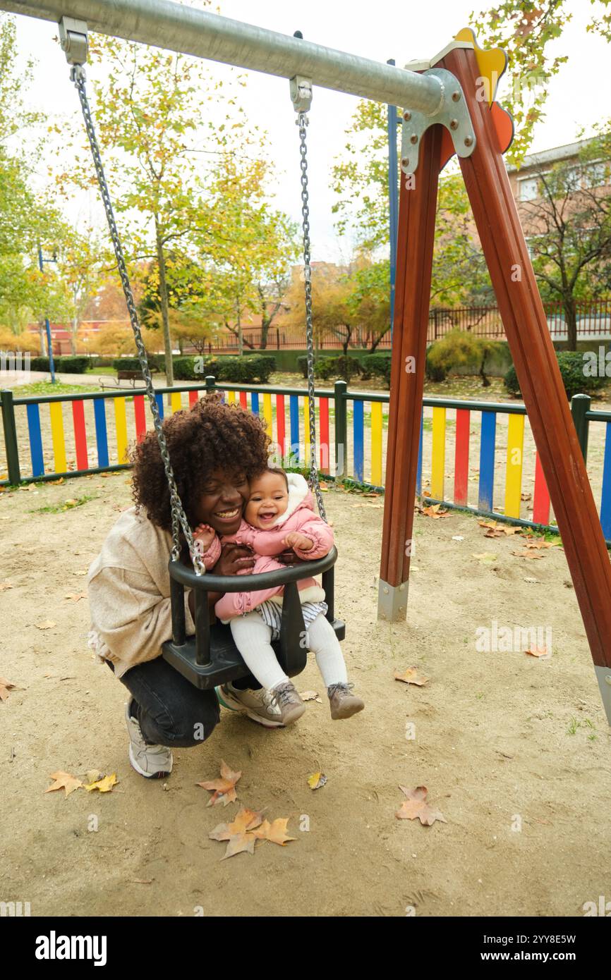 Mother pushing her laughing baby daughter on a swing in a playground ...