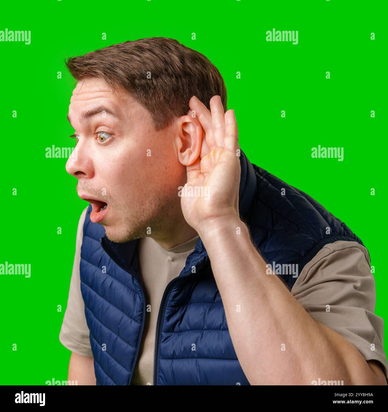 Surprised young man holding his hand to his ear, listening intently ...