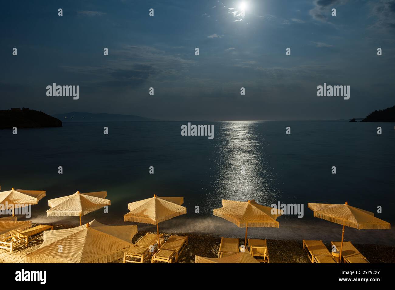 A peaceful moonlit beach with empty sun loungers and straw parasols ...