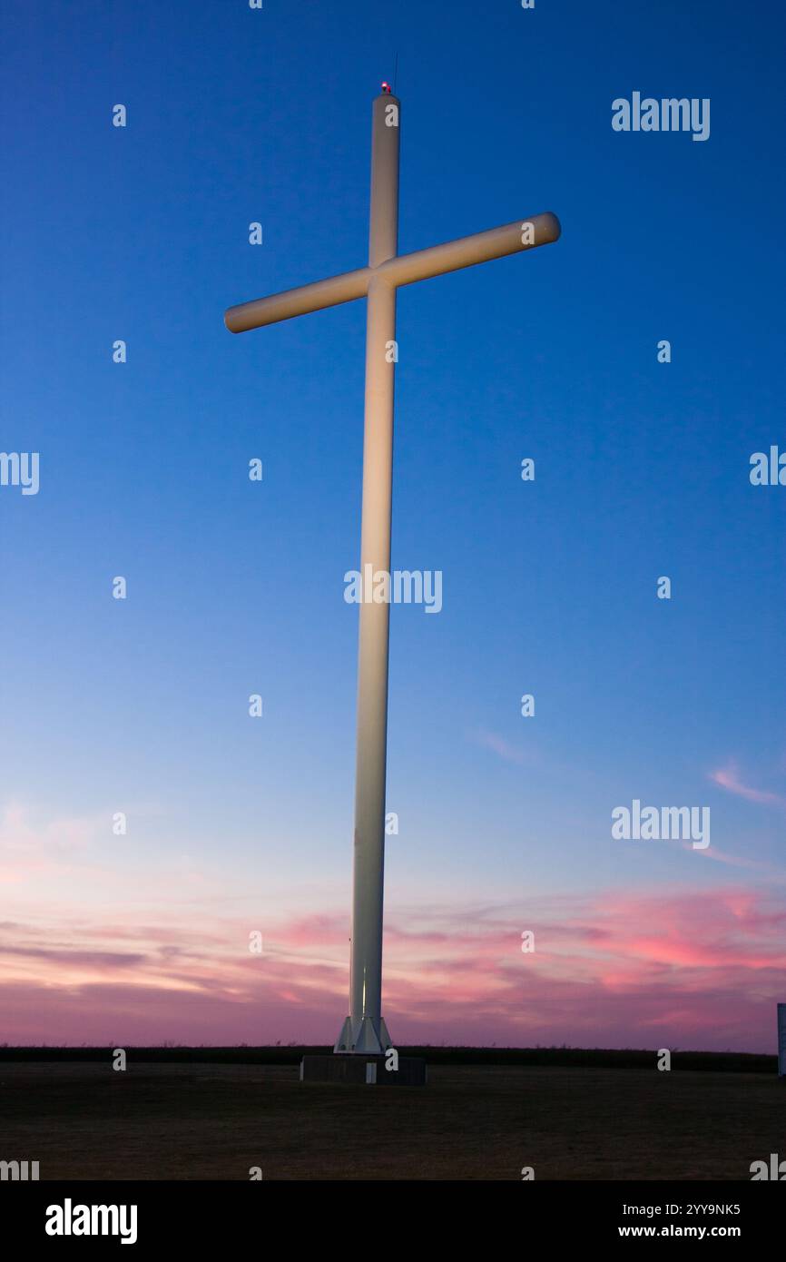 A tall white cross is lit up at night. The sky is blue and the sun is ...