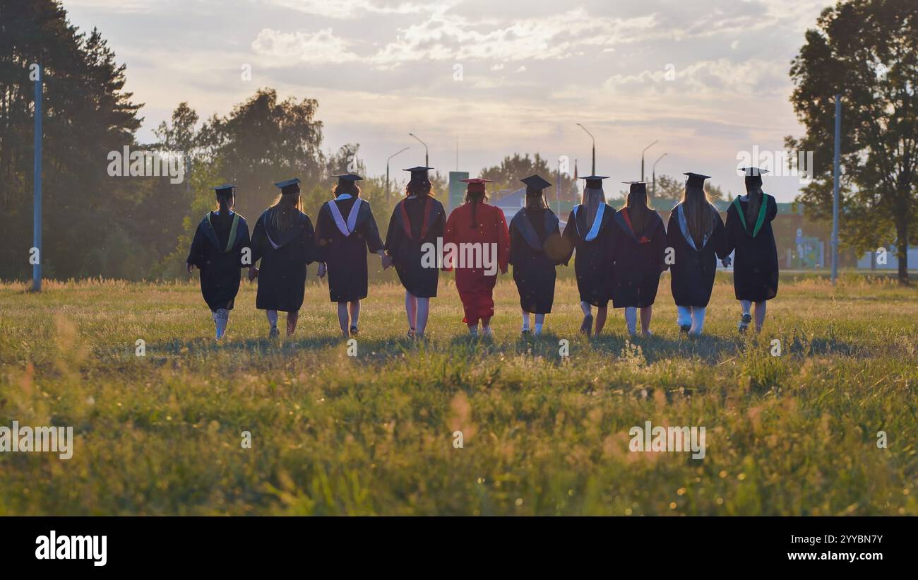 Ten college graduates walk across the field at sunset holding hands ...