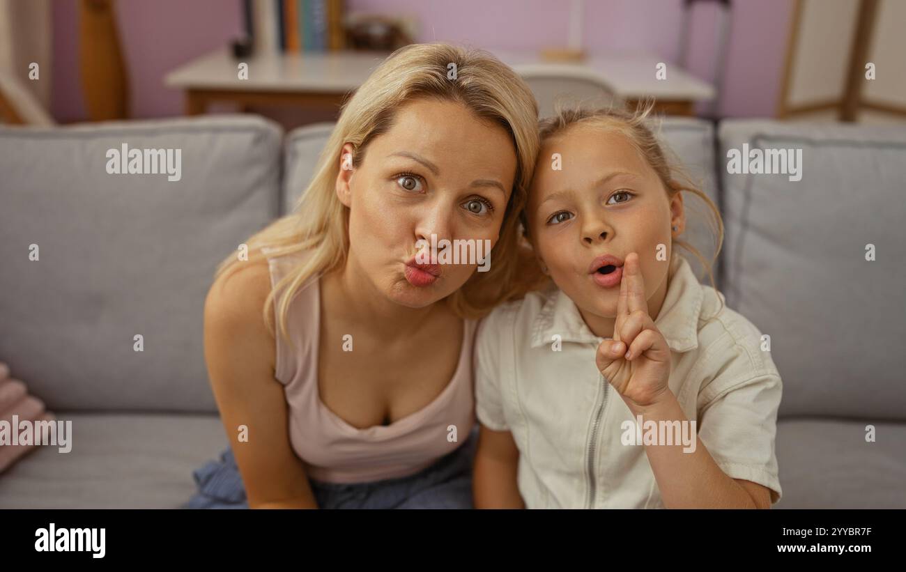 Blonde mother and daughter making funny faces together on a sofa in a cozy living room Stock ...