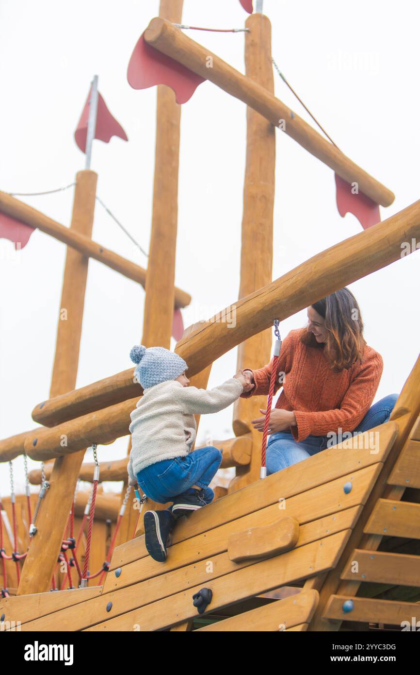 A mother assists her child climbing a wooden ship-themed playground ...
