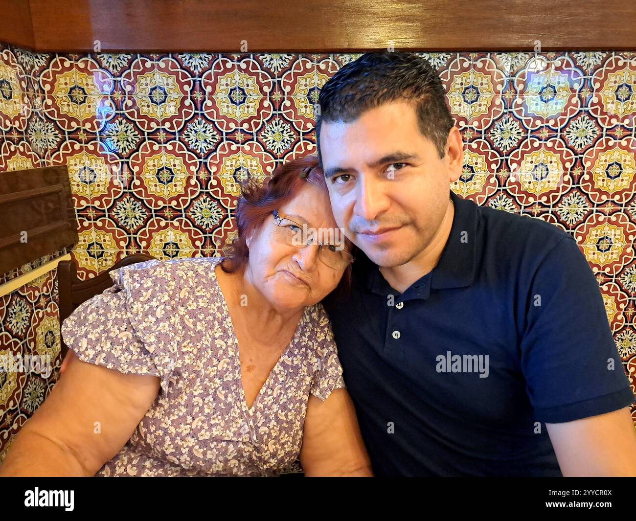 Elderly mother hugs her 40-year-old dark-skinned Latino son and they take a selfie photo to ...