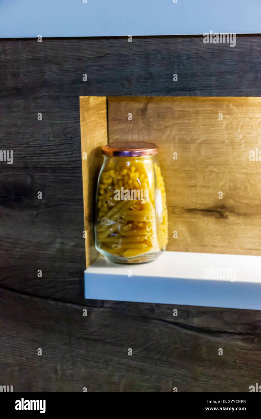 A glass jar filled with various types of pasta sits on a modern wooden ...
