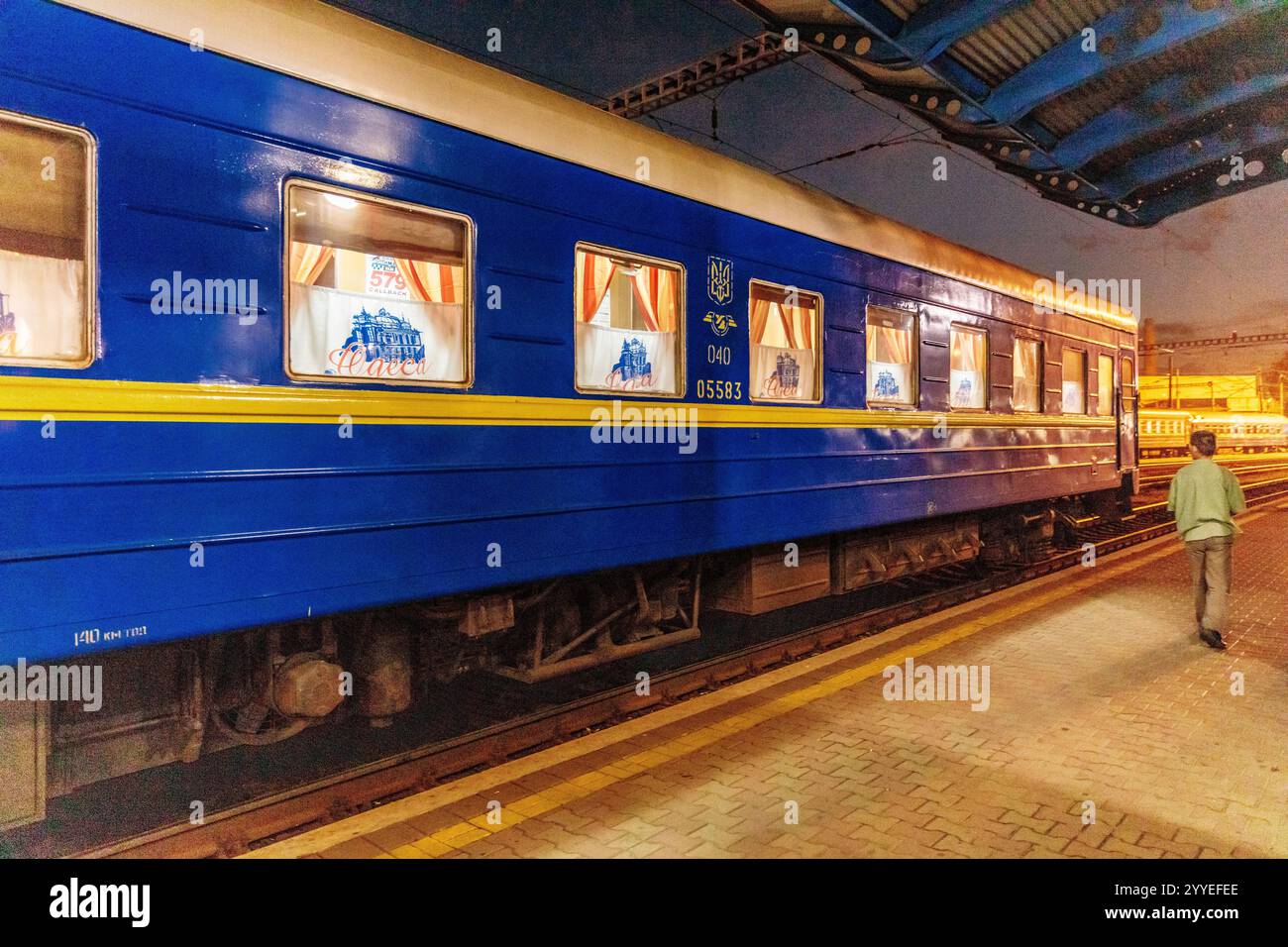 A blue train with a yellow stripe is parked at a station. A man is ...