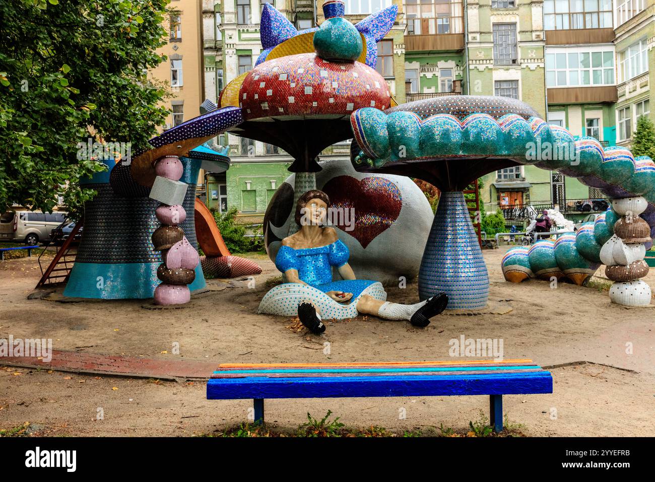 A woman statue sits on a bench in a park. The bench is blue and is next ...