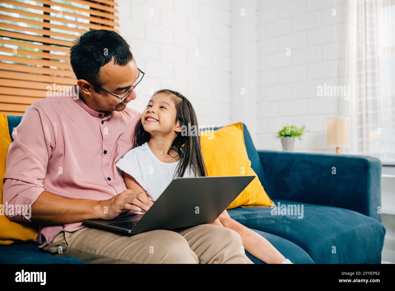 A smiling Asian father works from home on his laptop as his daughter ...