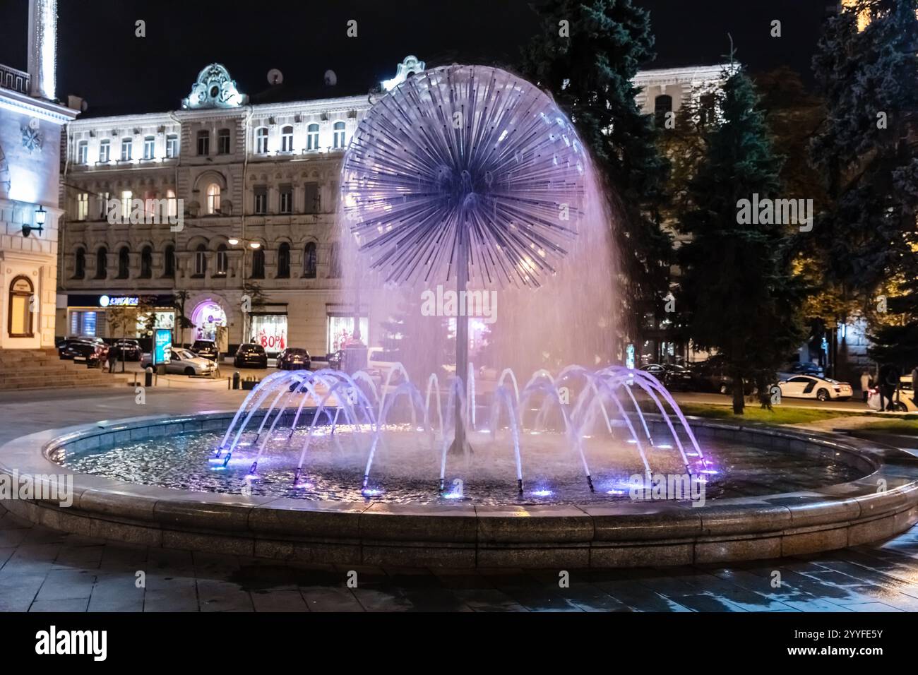A large fountain with a blue flower on top is lit up at night. The ...