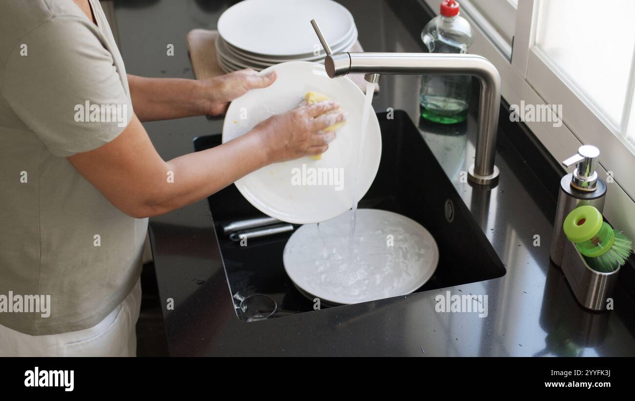 Woman washing and rinsing dishes by hand under a stream of water in the ...