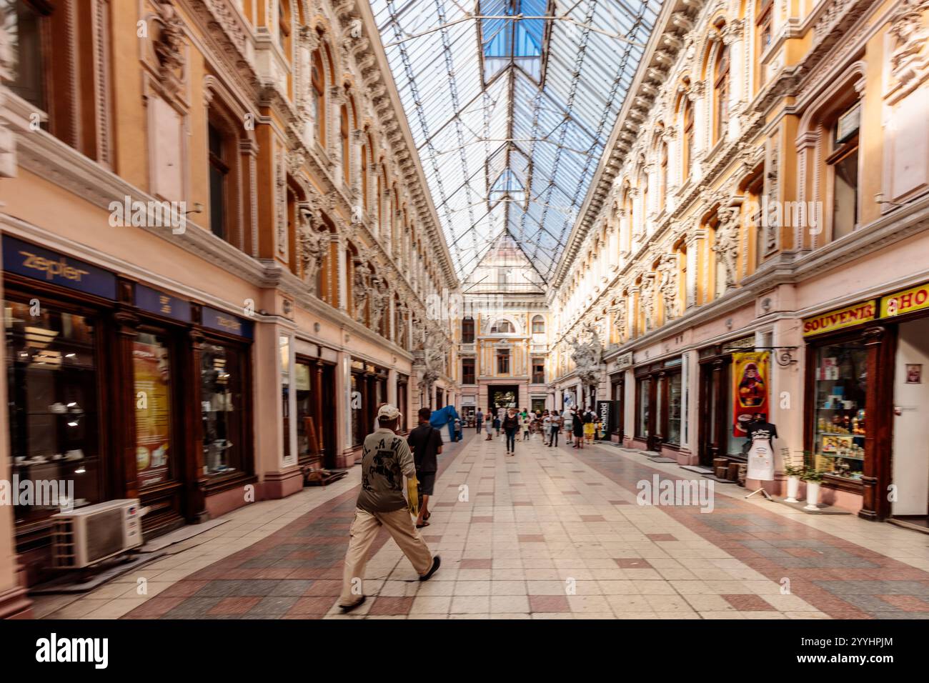 A long, narrow shopping mall with a blue roof. People are walking ...