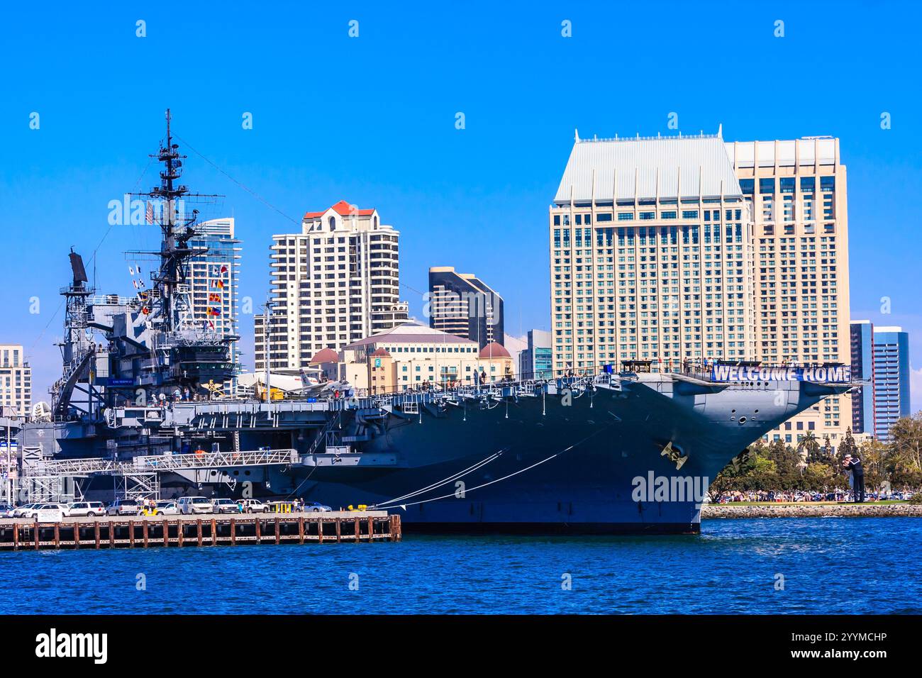 A large navy ship is docked in front of a large hotel. The ship is the ...