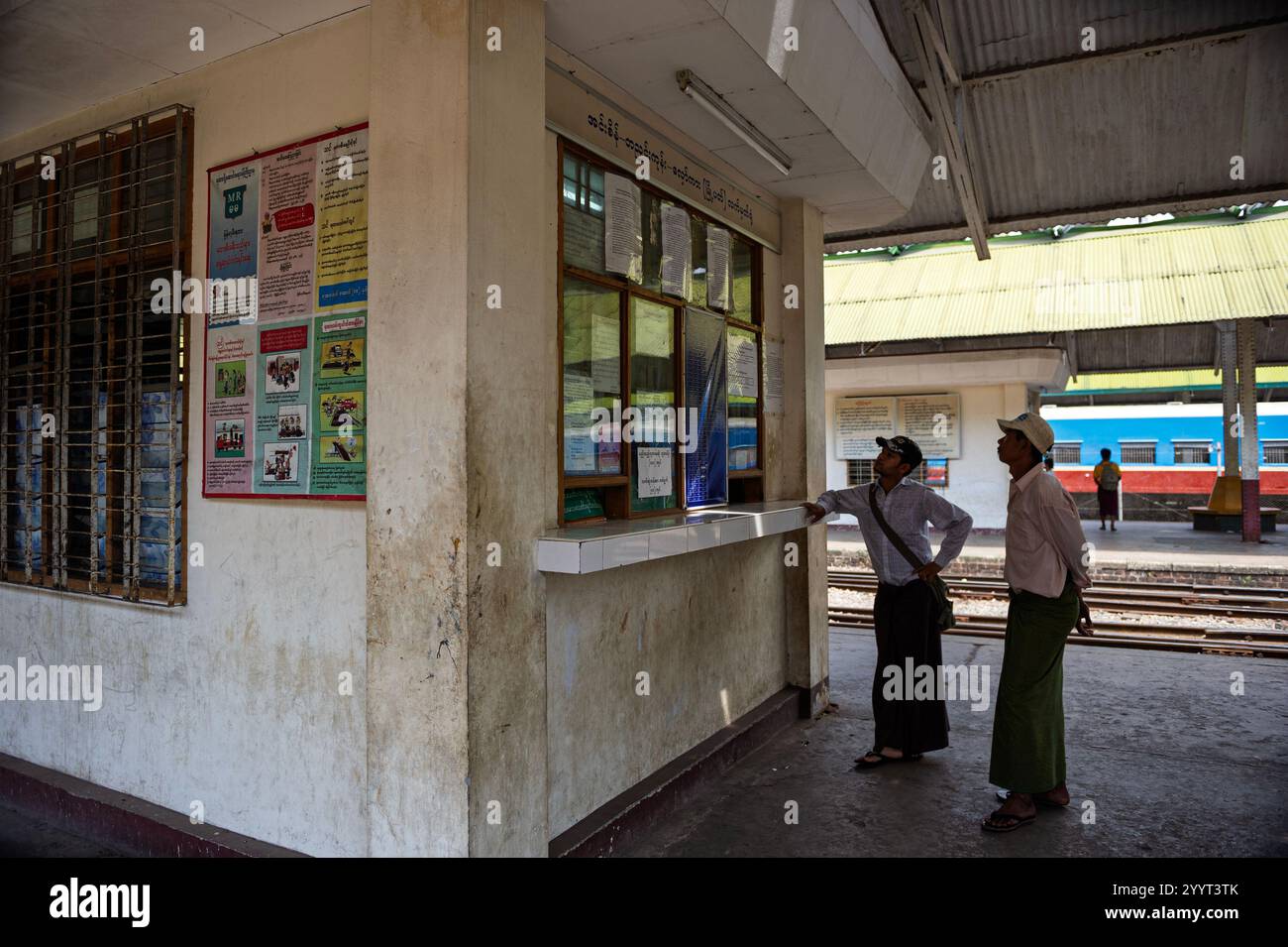 Two men standing at a train station ticket counter examining timetables ...